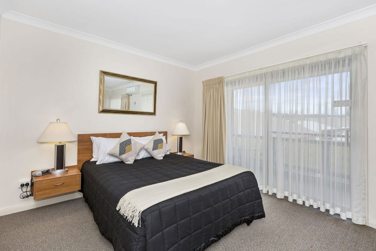 Quarto de hotel com cama, duas luminárias de cabeceira, mesa de apoio, espelho e uma janela grande com cortinas leves.
