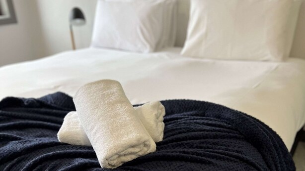 Cama bem arrumada com lençóis brancos e um cobertor azul escuro, um travesseiro branco e uma luminária de cabeceira.
