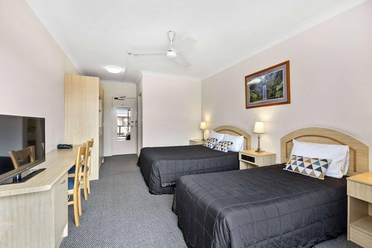 Quarto de hotel com duas camas, uma escrivaninha, televisão e ventoinha no teto.
