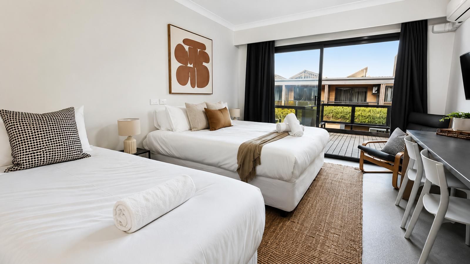 Quarto de hotel com duas camas, uma escrivaninha e vista para um edifício através de grandes janelas.