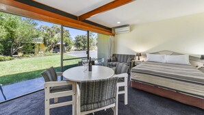 Quarto moderno com uma janela grande, uma mesa redonda com cadeiras, uma cama e vista para um gramado verde e árvores.