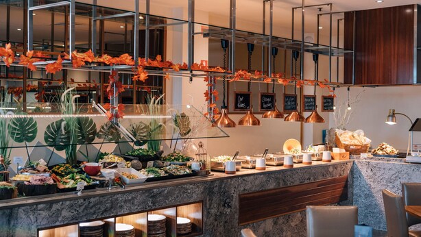Uma área de buffet com diversos alimentos, um espaço de refeições com cadeiras e uma decoração com plantas.