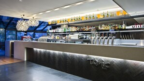 Uma área de bar com piso em padrão herringbone, um balcão com vitrine de vidro e diversos frascos e copos nas prateleiras.