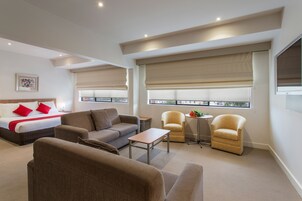 Quarto de hotel moderno com cama, sofá, poltronas, uma mesa pequena e grandes janelas com persianas.