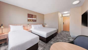 Quarto de hotel com duas camas, uma escrivaninha, uma cadeira, uma televisão e uma porta que dá para outro cômodo.