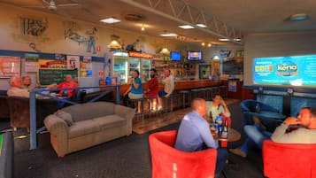 Espaço de bar com assentos, mesa de bilhar e televisão exibindo uma partida.