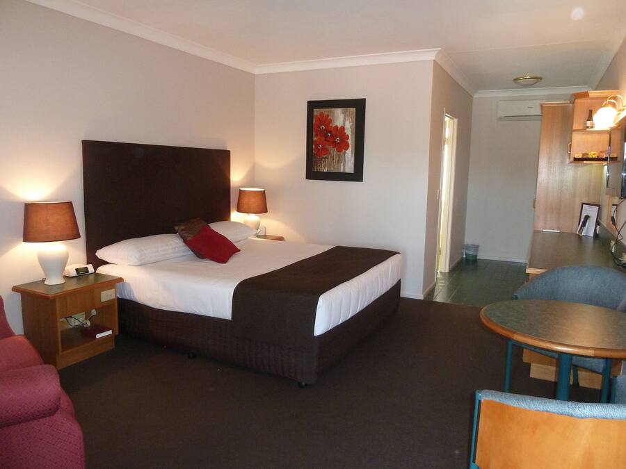 Quarto de hotel com cama, duas luminárias de cabeceira, travesseiro vermelho, mesa de cabeceira de madeira, uma pequena mesa e uma cadeira.