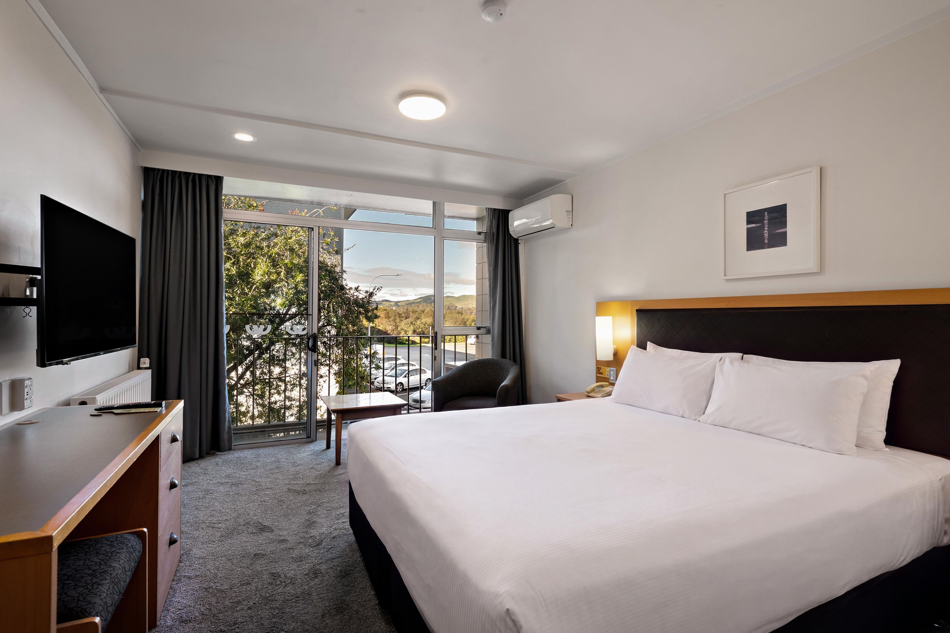 Quarto de hotel com uma cama grande, uma escrivaninha com televisão, uma cadeira e vista para o exterior.
