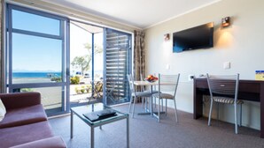 Quarto de hotel com grande porta de vidro deslizante para a varanda, televisão de tela plana na parede, sofá, mesa de jantar com cadeiras e uma pequena escrivaninha.