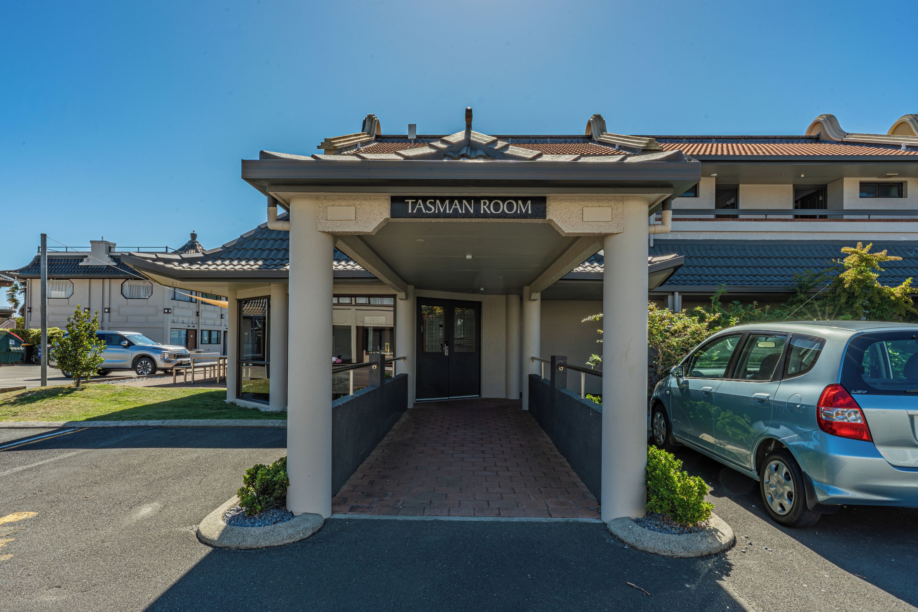 Entrada de um edifício com uma placa indicando 'TASMAN ROOM', um caminho pavimentado que leva à porta e um carro estacionado à direita.