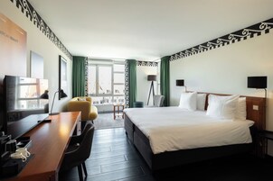 Quarto de hotel com cama, escrivaninha, cadeira, luminária e vista para a paisagem urbana pela janela.