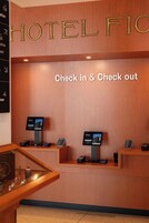 Uma área de recepção de hotel com um balcão de madeira, dois totens de check-in e uma placa com a inscrição 'HOTEL FIC'.