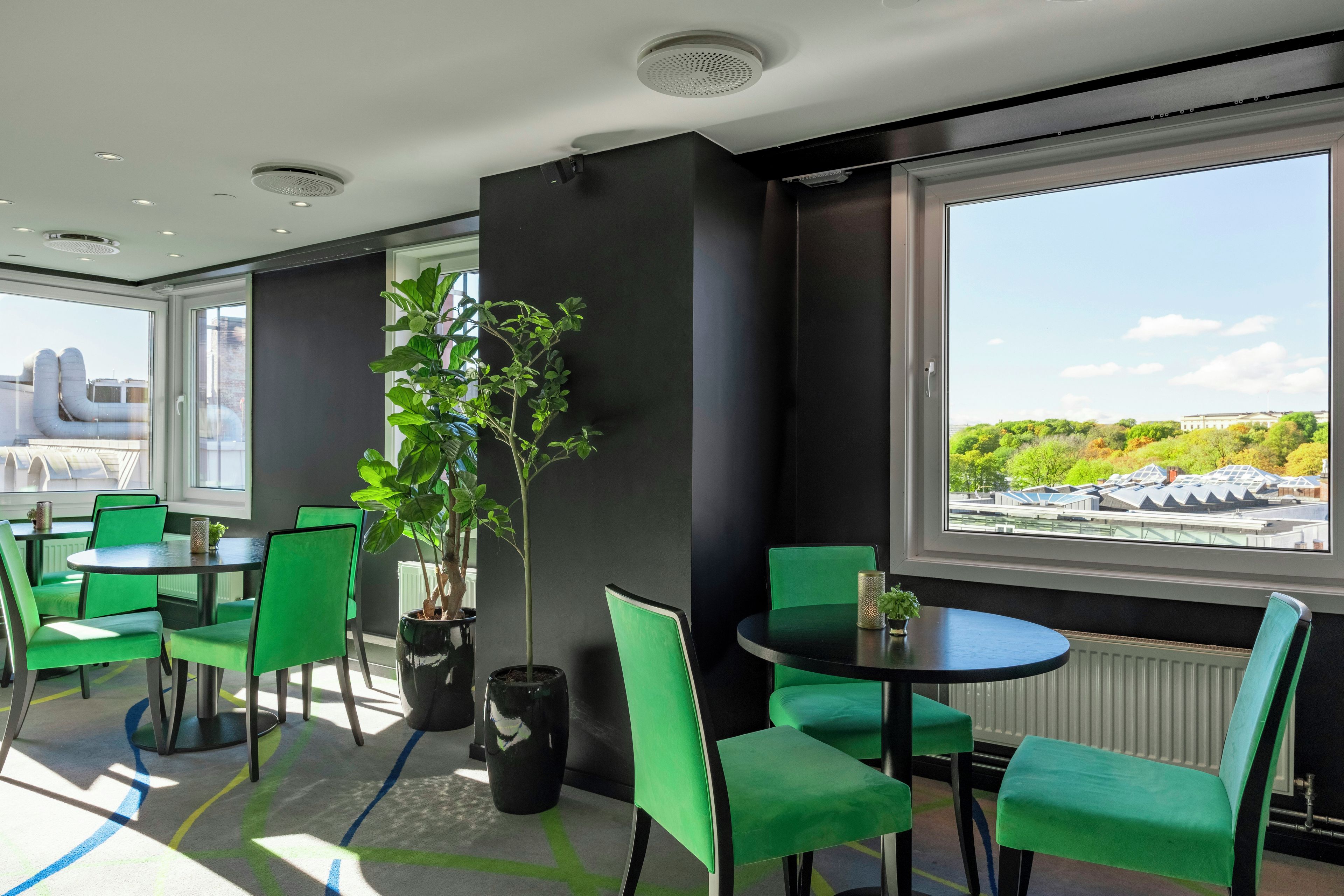 Decoração moderna com cadeiras verdes, planta em vaso e uma grande janela com vista para edifícios e áreas verdes.
