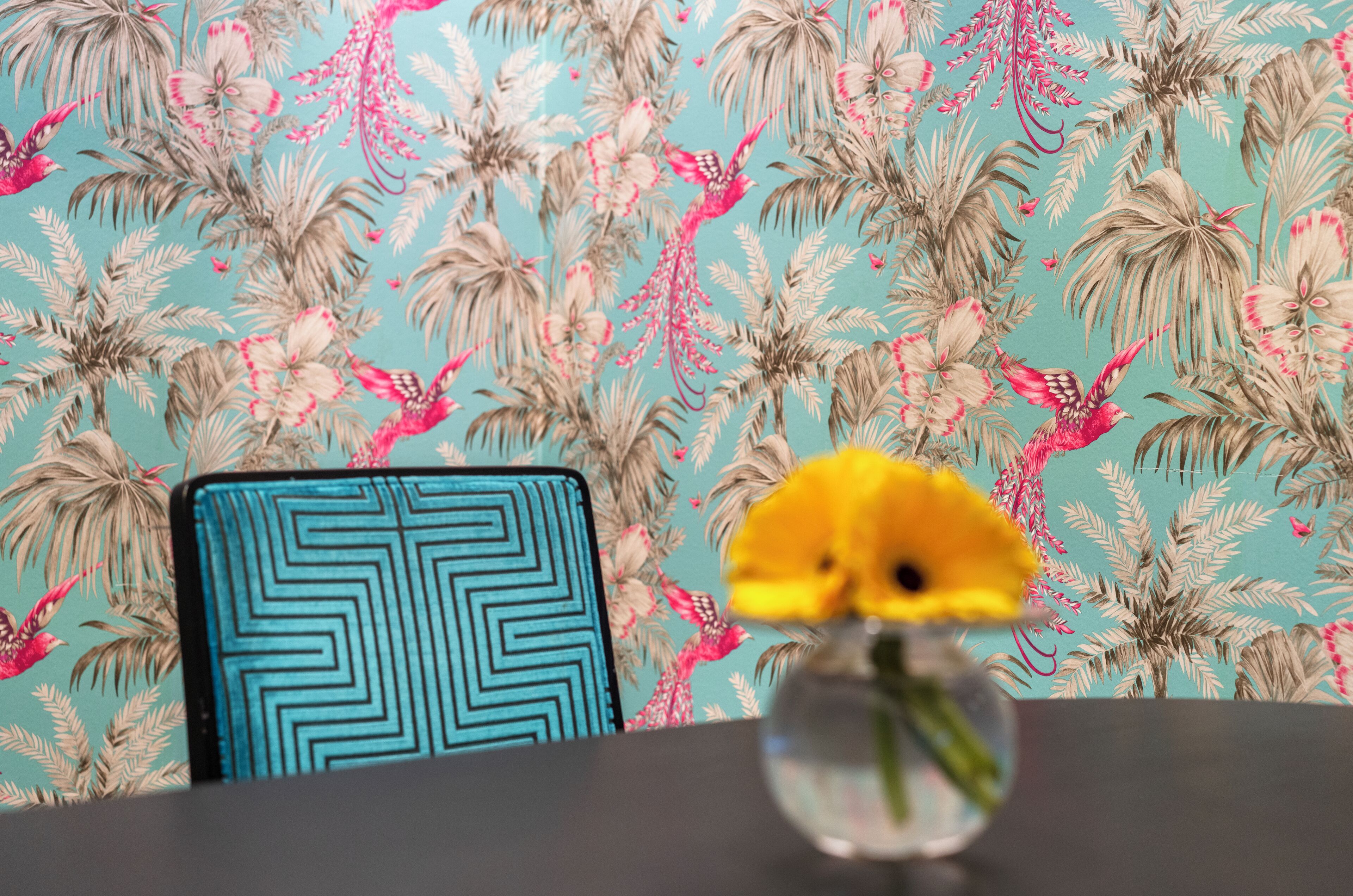 Papel de parede estampado com desenhos florais e geométricos, uma cadeira azul com padrão em zigue-zague e um vaso com uma flor amarela sobre uma mesa.
