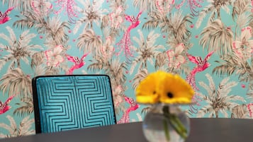 Papel de parede estampado com desenhos florais e geométricos, uma cadeira azul com padrão em zigue-zague e um vaso com uma flor amarela sobre uma mesa.