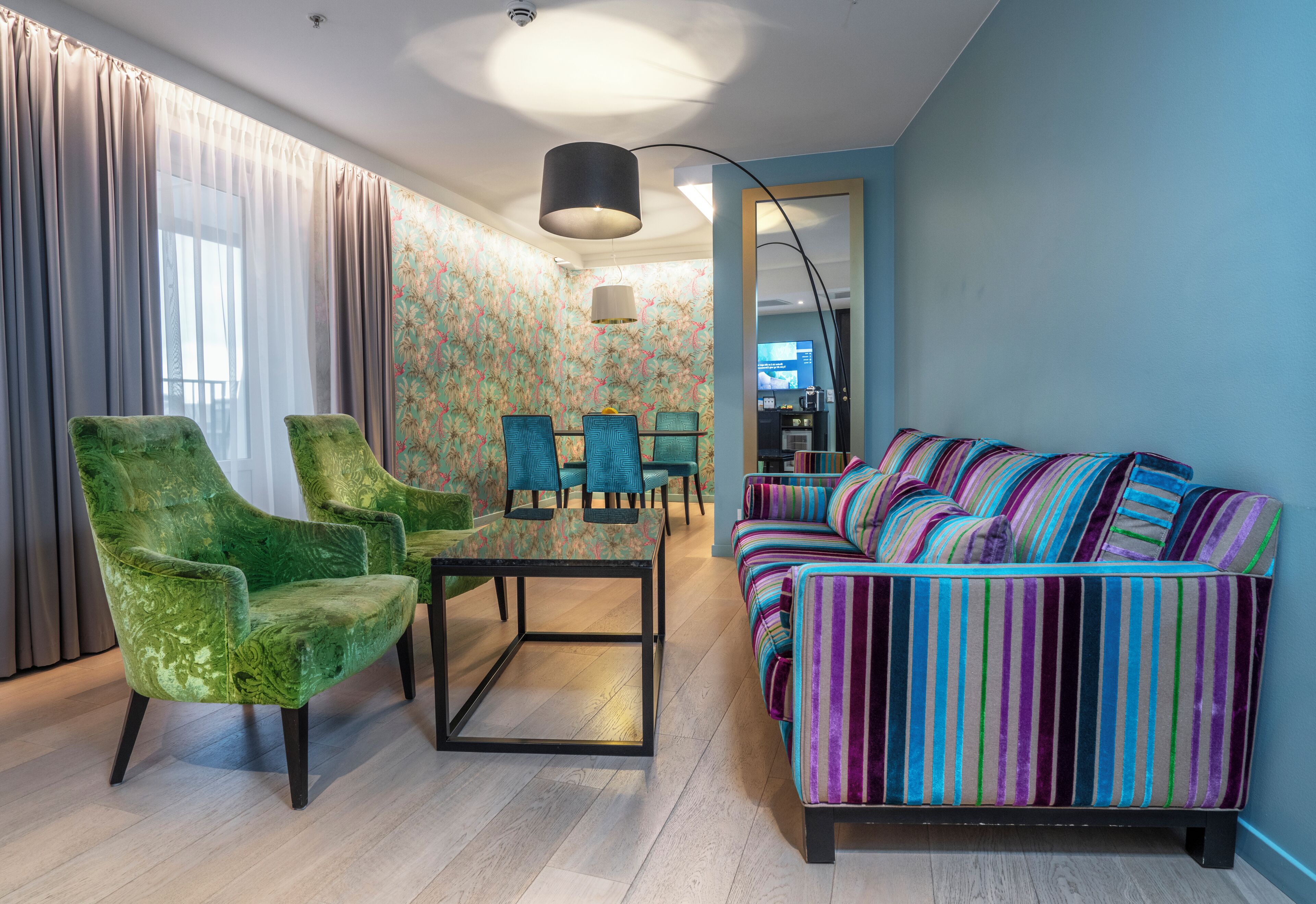 Uma sala de estar com poltronas de veludo verde, um sofá riscado, uma mesa de centro com tampo de vidro e um espaço de jantar com cadeiras azul-turquesa.