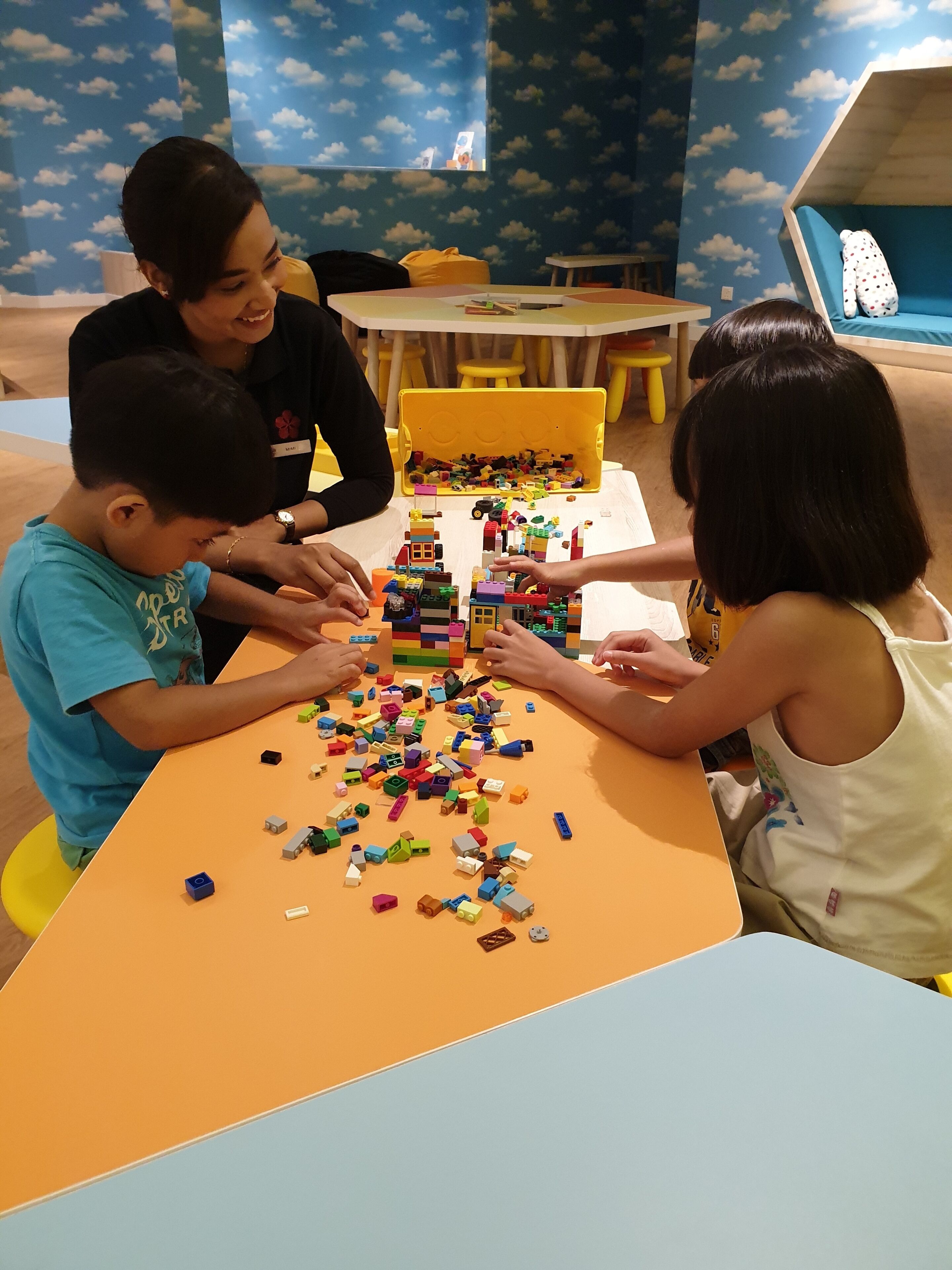 Um grupo de pessoas brincando com blocos de construção em uma mesa, em um quarto com parede azul e padrão de nuvens.