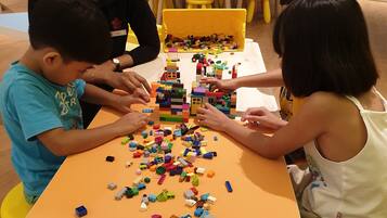 Um grupo de pessoas brincando com blocos de construção em uma mesa, em um quarto com parede azul e padrão de nuvens.