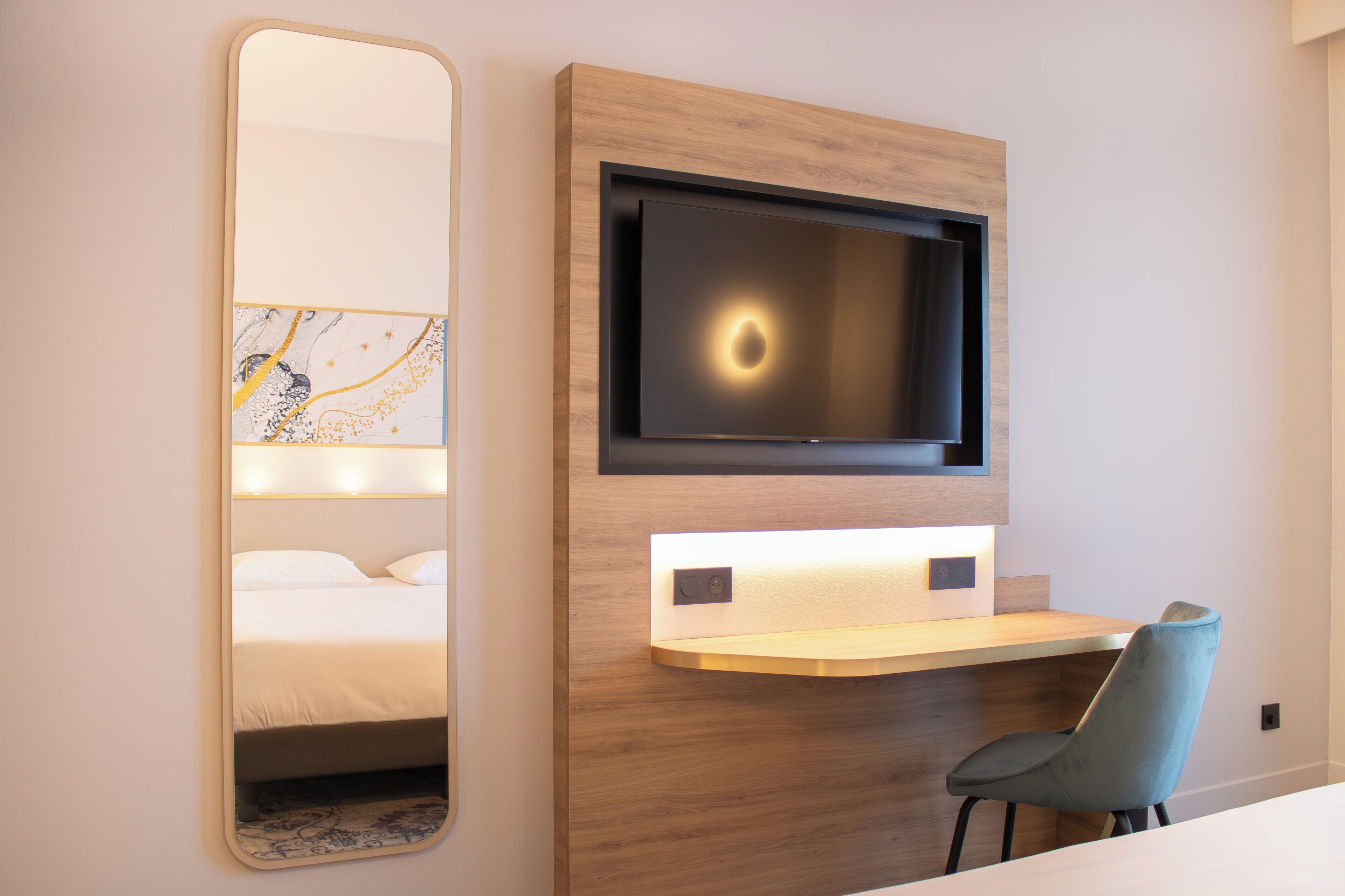 Quarto de hotel moderno com uma cama grande, uma escrivaninha com cadeira, televisão fixada na parede e espelho também fixado na parede.