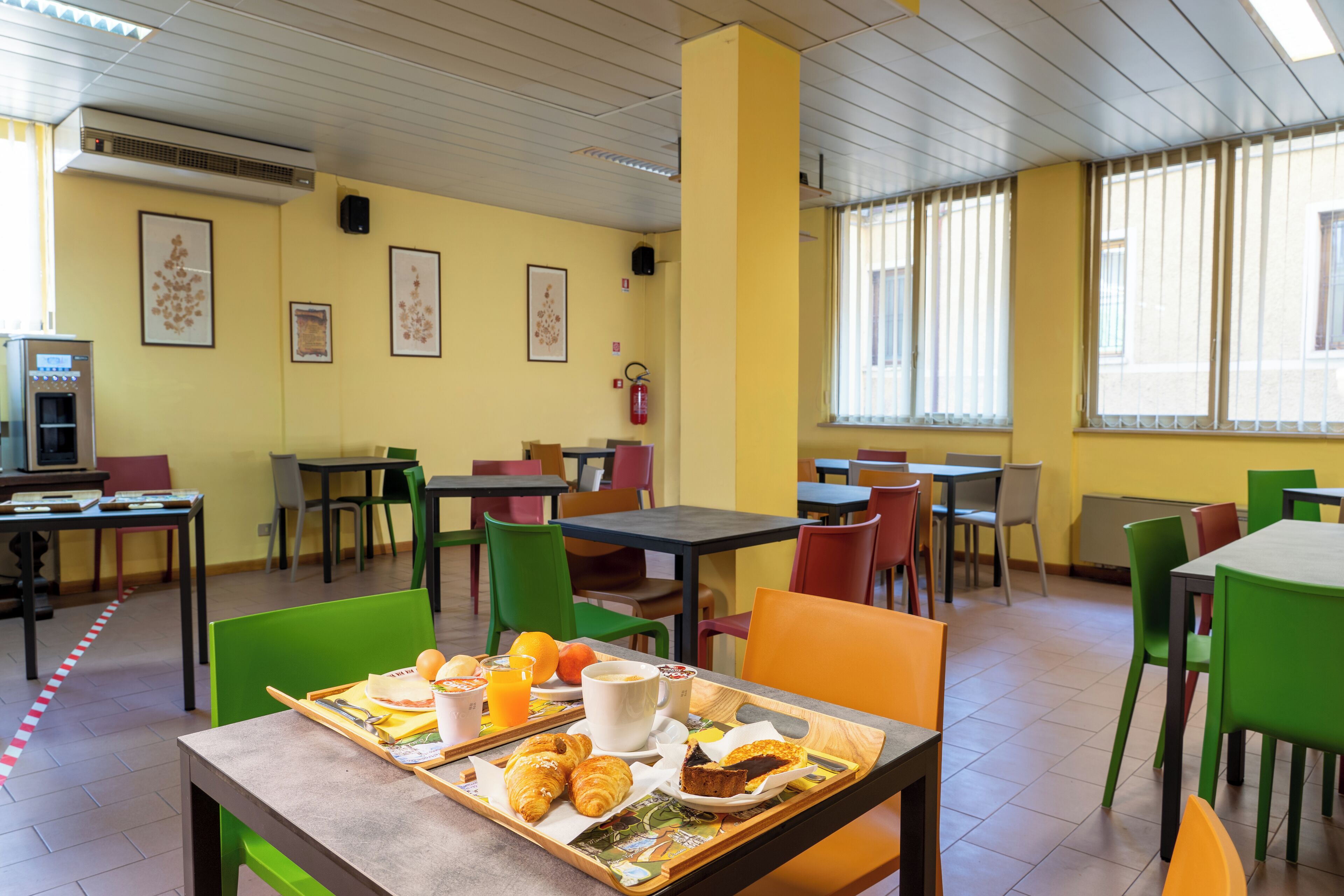 Uma área de refeições com mesas e cadeiras, um tabuleiro com comida e bebidas, e um extintor de incêndio na parede.