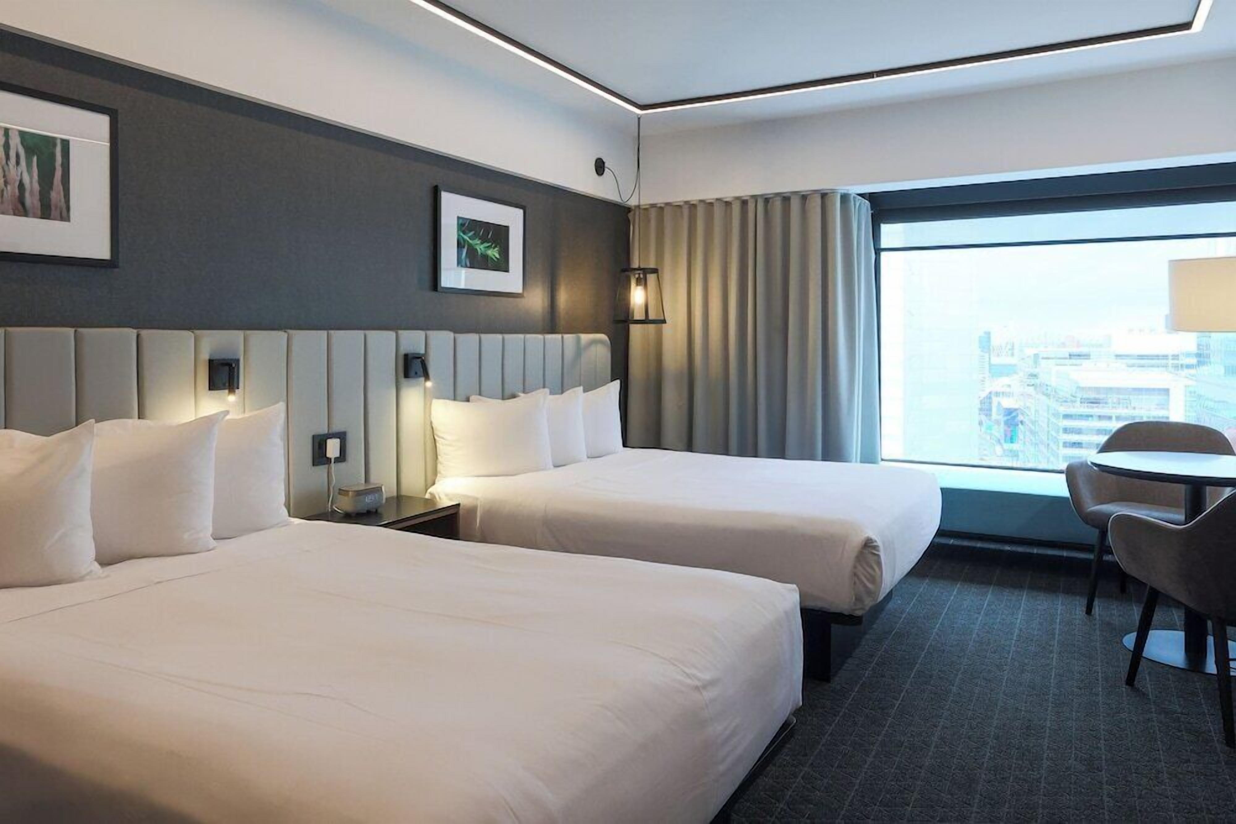 Quarto de hotel com duas camas, uma escrivaninha, uma cadeira, vista para a cidade e um quadro na parede.