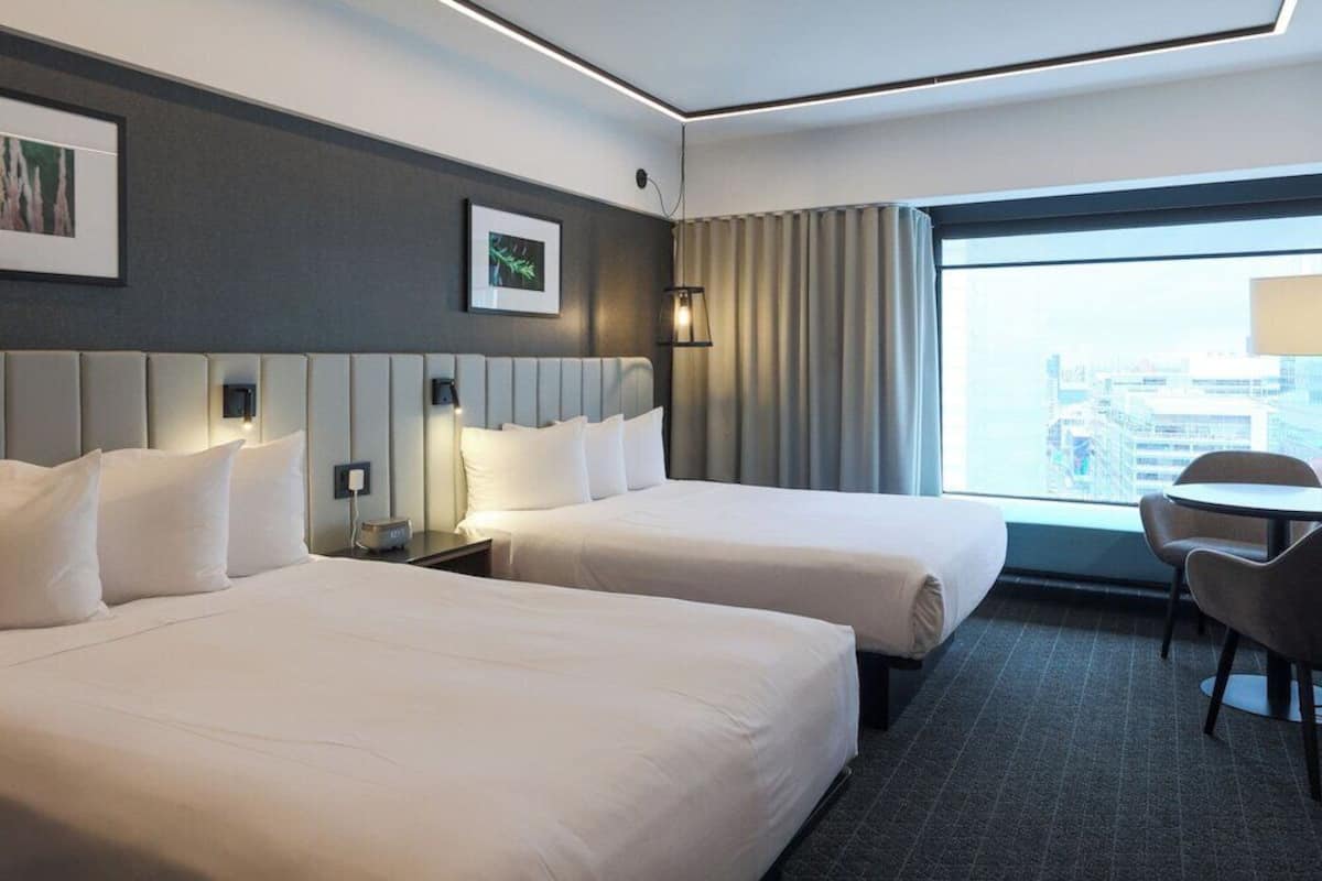Quarto de hotel com duas camas, uma escrivaninha, uma cadeira, vista para a cidade e um quadro na parede.