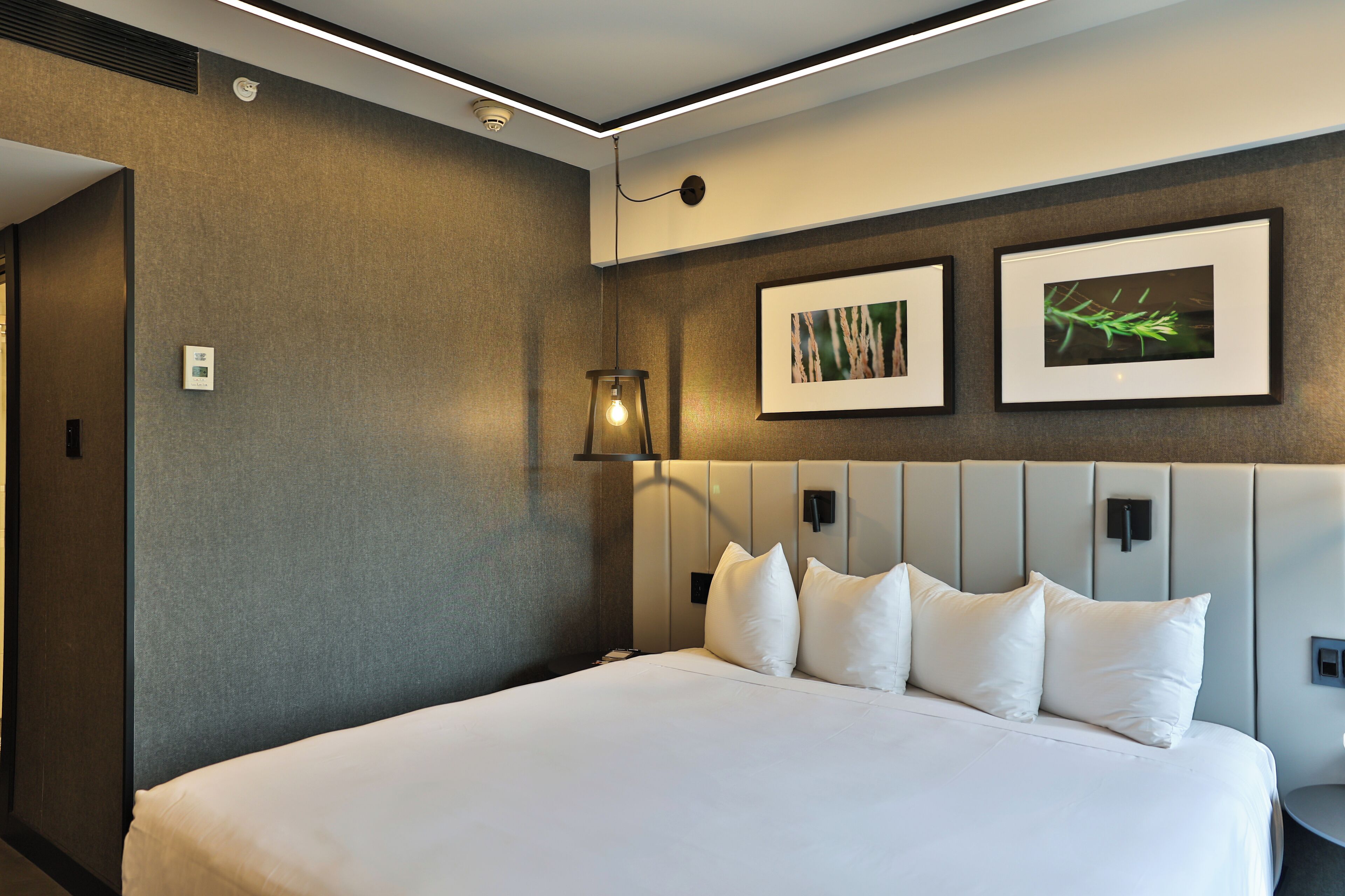Quarto de hotel moderno com uma cama grande, cabeceira com várias almofadas, uma luz fixada na parede e duas imagens emolduradas.