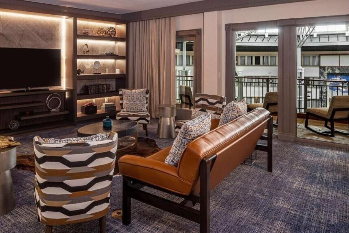 Quarto de hotel moderno com uma grande televisão de tela plana, um sofá, cadeiras estampadas, uma mesa de centro e uma estante.