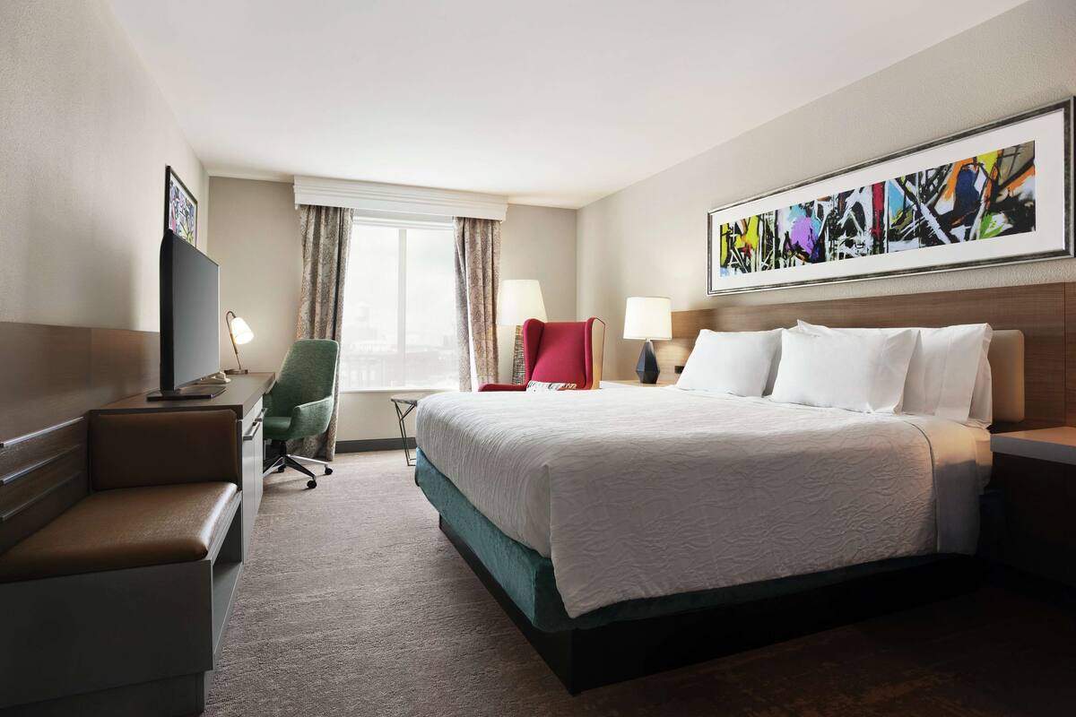 Quarto de hotel com cama, escrivaninha com computador, cadeira, televisão e um quadro na parede.