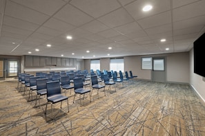 Sala de conferências com fileiras de cadeiras, piso carpetado e teto com iluminação embutida.