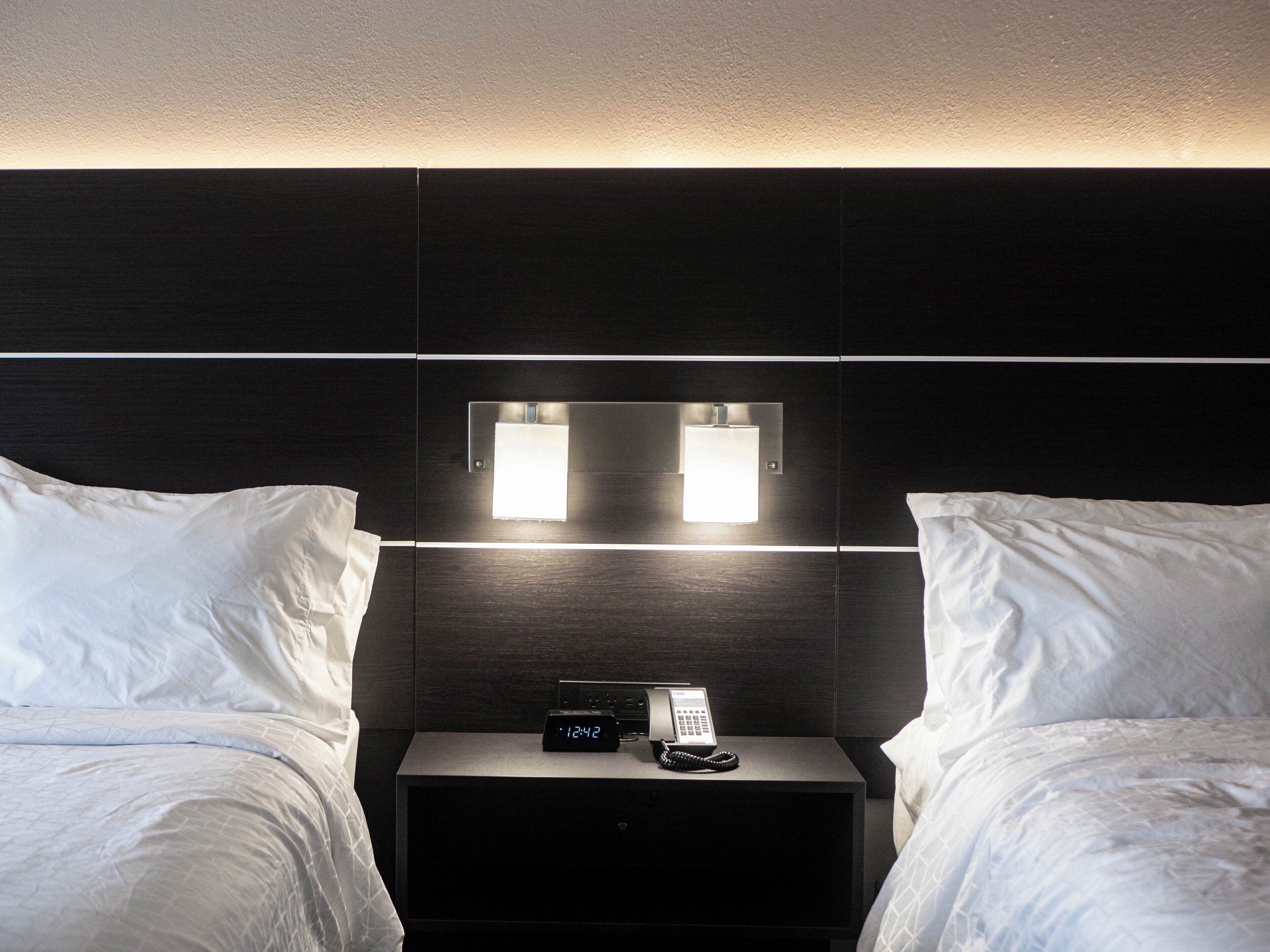 Quarto de hotel com duas camas, uma mesa de cabeceira com telefone e relógio digital, e iluminação fixada na parede.