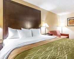 Quarto de hotel com uma cama grande, cabeceira, mesa de cabeceira com luminária, cadeira e um quadro na parede.