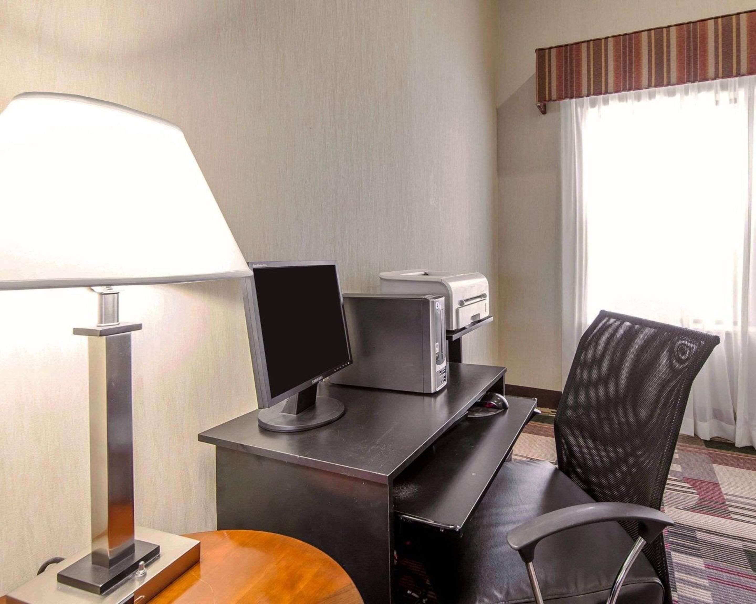 Quarto de hotel com escrivaninha, monitor de computador, impressora, luminária e cadeira.
