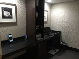 Quarto de hotel com escrivaninha, computador, cadeira e uma moldura na parede.