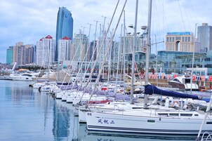Uma marina com vários barcos à vela atracados, um horizonte urbano ao fundo e um céu claro.