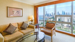 Quarto de hotel com uma janela grande que oferece vista para a cidade, um sofá com almofadas estampadas, uma mesa de centro de vidro e um quadro na parede.