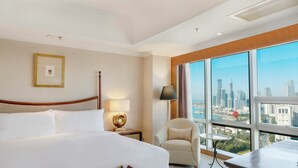Quarto de hotel com uma cama grande, uma cadeira, uma mesa e vista para a paisagem urbana através de janelas amplas.