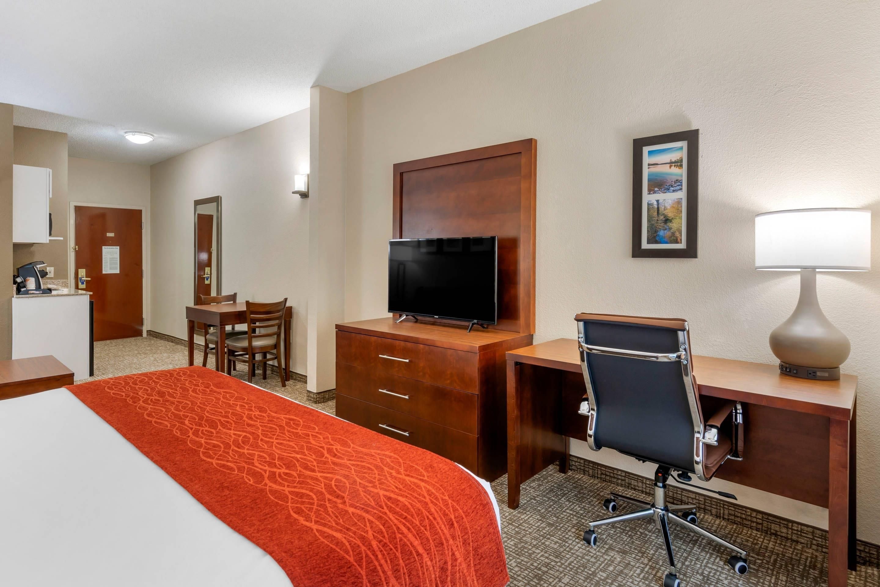 Quarto de hotel com cama, escrivaninha, cadeira, televisão, luminária e quadro pendurado na parede.
