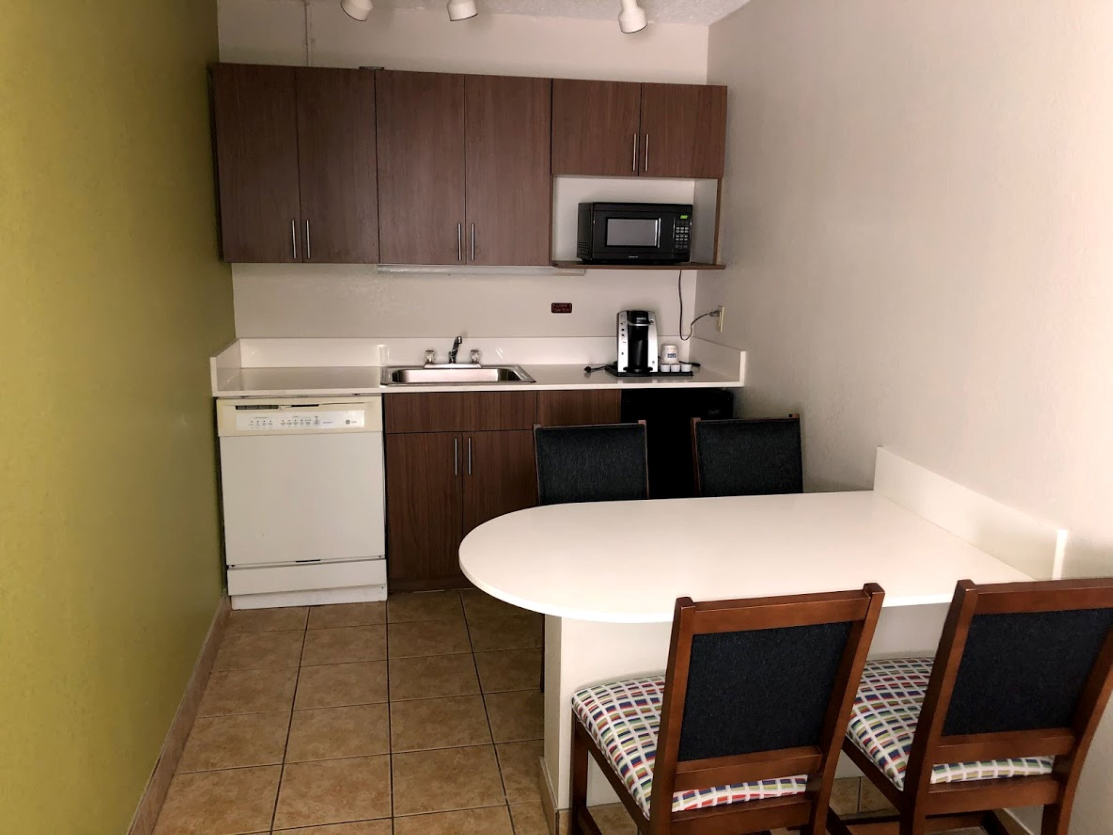 Cozinha compacta equipada com micro-ondas, cafeteira e máquina de lavar loiça. Inclui uma mesa de jantar com duas cadeiras.