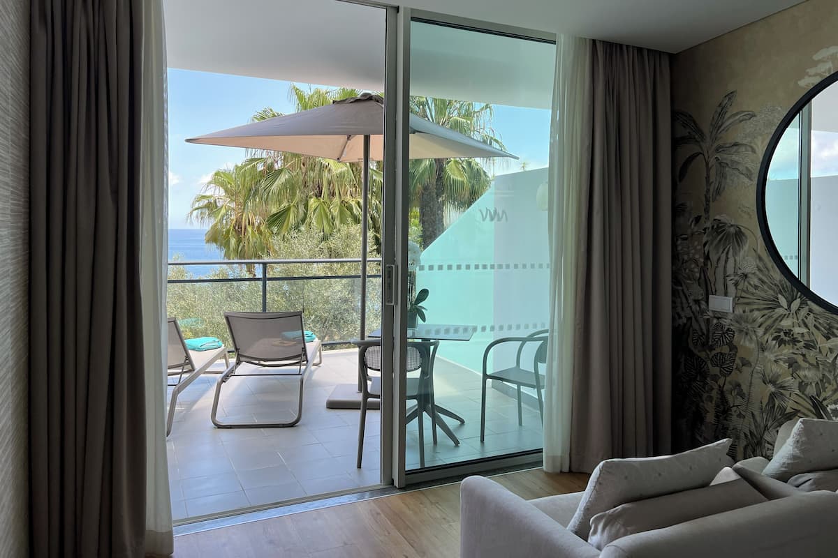 Quarto de hotel com varanda, área de refeições e vista para palmeiras e o mar.