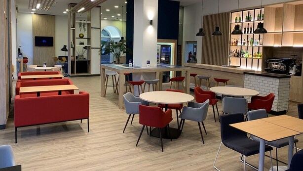 Um café moderno com várias áreas de estar, incluindo cadeiras vermelhas e cinzentas, mesas de madeira e um bar com prateleiras cheias de garrafas.