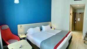 Quarto de hotel com uma cama, uma cadeira vermelha, uma mesinha e um criado-mudo.