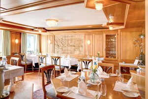 Uma sala de jantar formal com lambris de madeira, lustres e um grande mural. Mesas com toalhas brancas e porcelana fina.