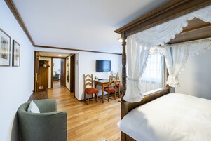 Quarto de hotel com piso de madeira, cama com roupa de cama branca, escrivaninha com cadeiras, televisão e corredor que leva a outro quarto.