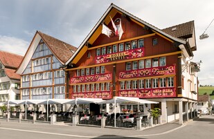 Um hotel com estilo arquitetônico europeu tradicional, destacando uma placa de identificação e uma área externa com mesas e cadeiras.