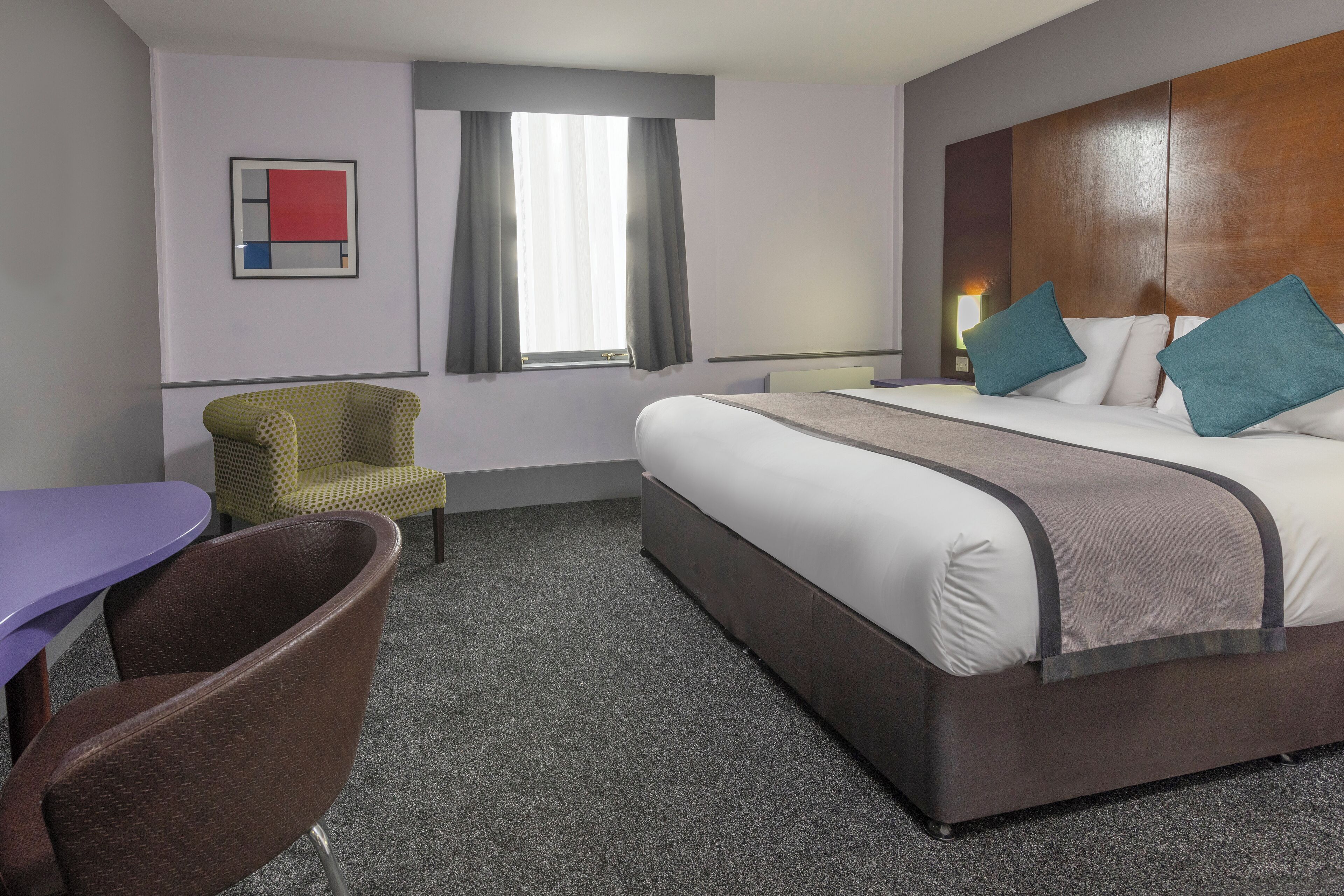Quarto de hotel com uma cama grande, uma mesinha, uma cadeira e uma janela com cortinas.