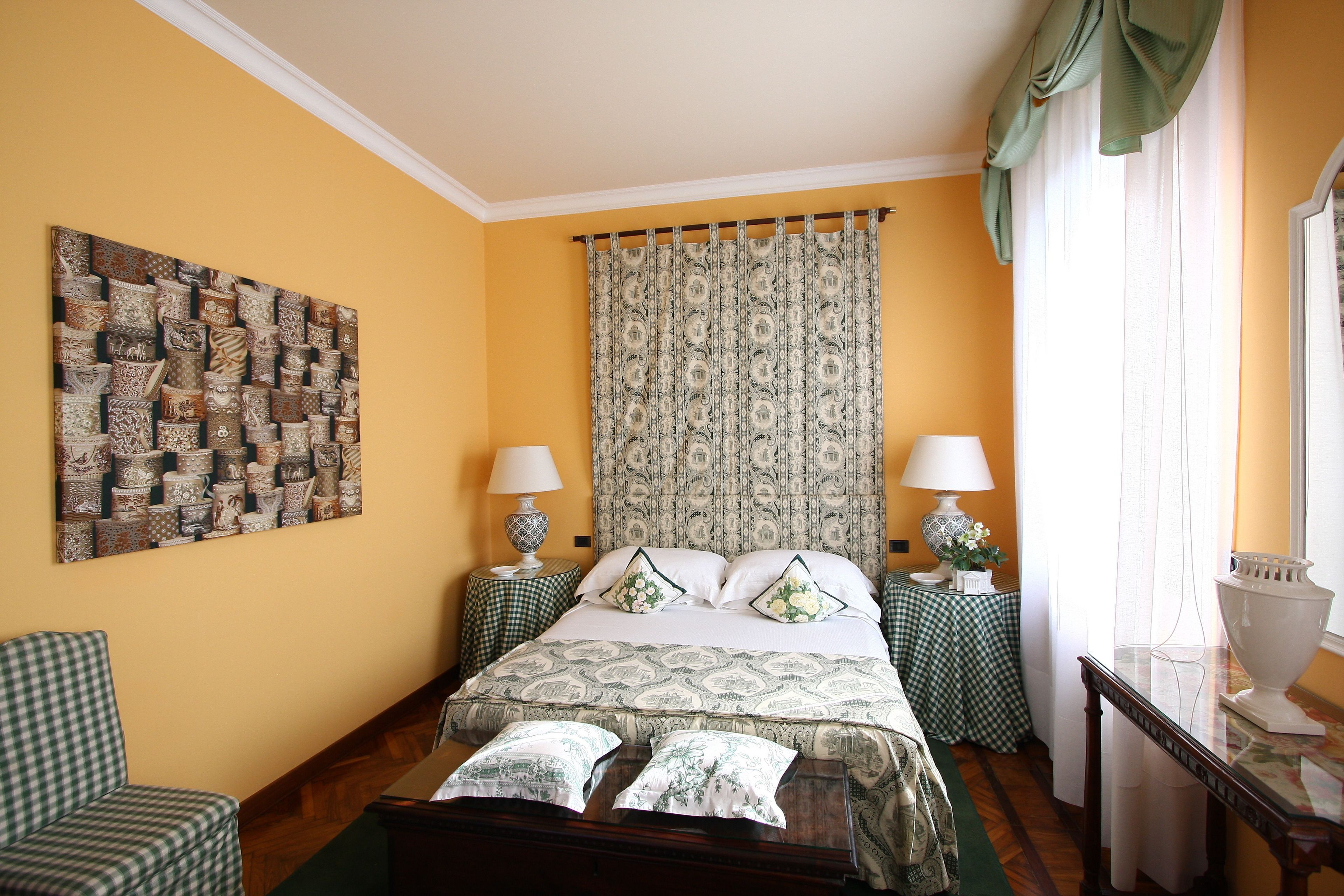 Quarto de hotel com uma cama, duas mesas de cabeceira, uma cadeira, um objeto decorativo na parede e uma janela com cortinas.