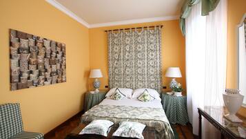 Quarto de hotel com uma cama, duas mesas de cabeceira, uma cadeira, um objeto decorativo na parede e uma janela com cortinas.