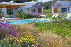 Uma área à beira da piscina com espreguiçadeiras, um pérgola e um pequeno jardim com flores roxas e amarelas.
