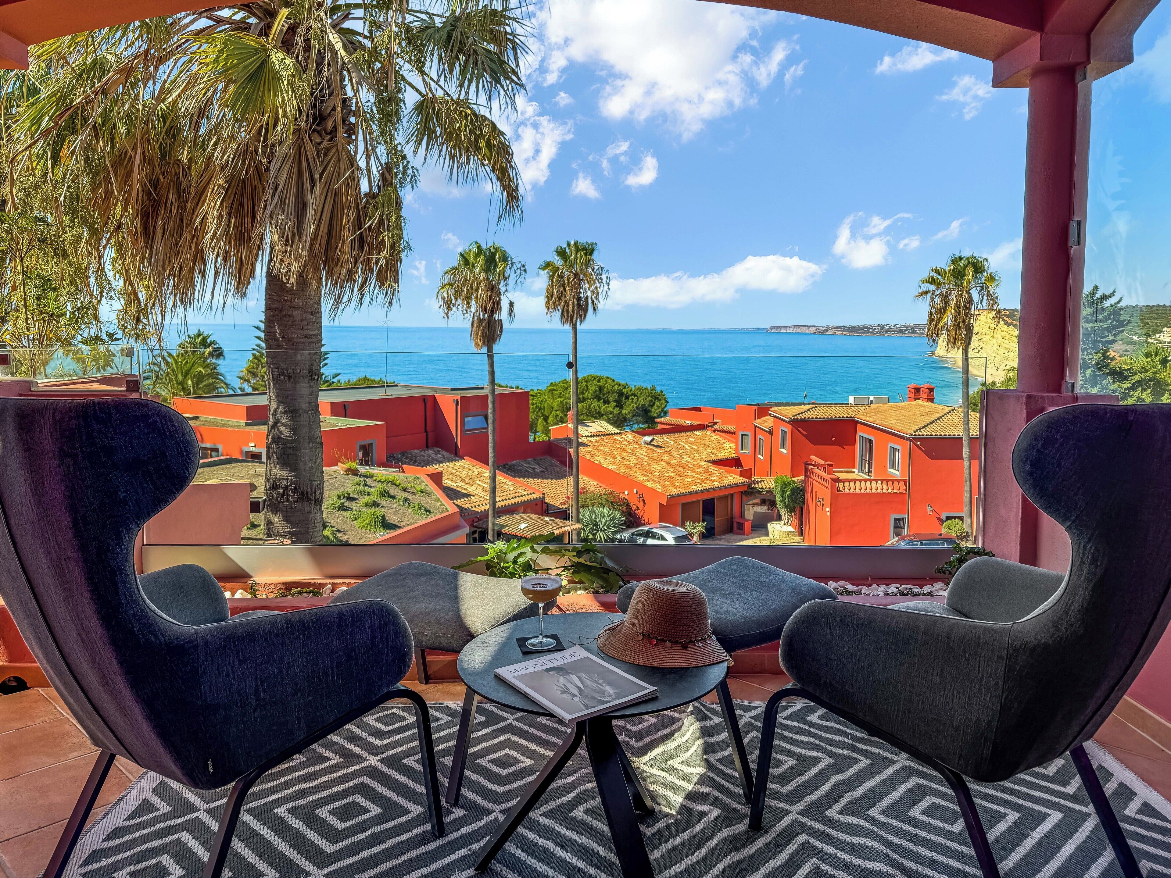 Uma varanda com duas cadeiras, uma mesa e vista para o mar e edifícios.